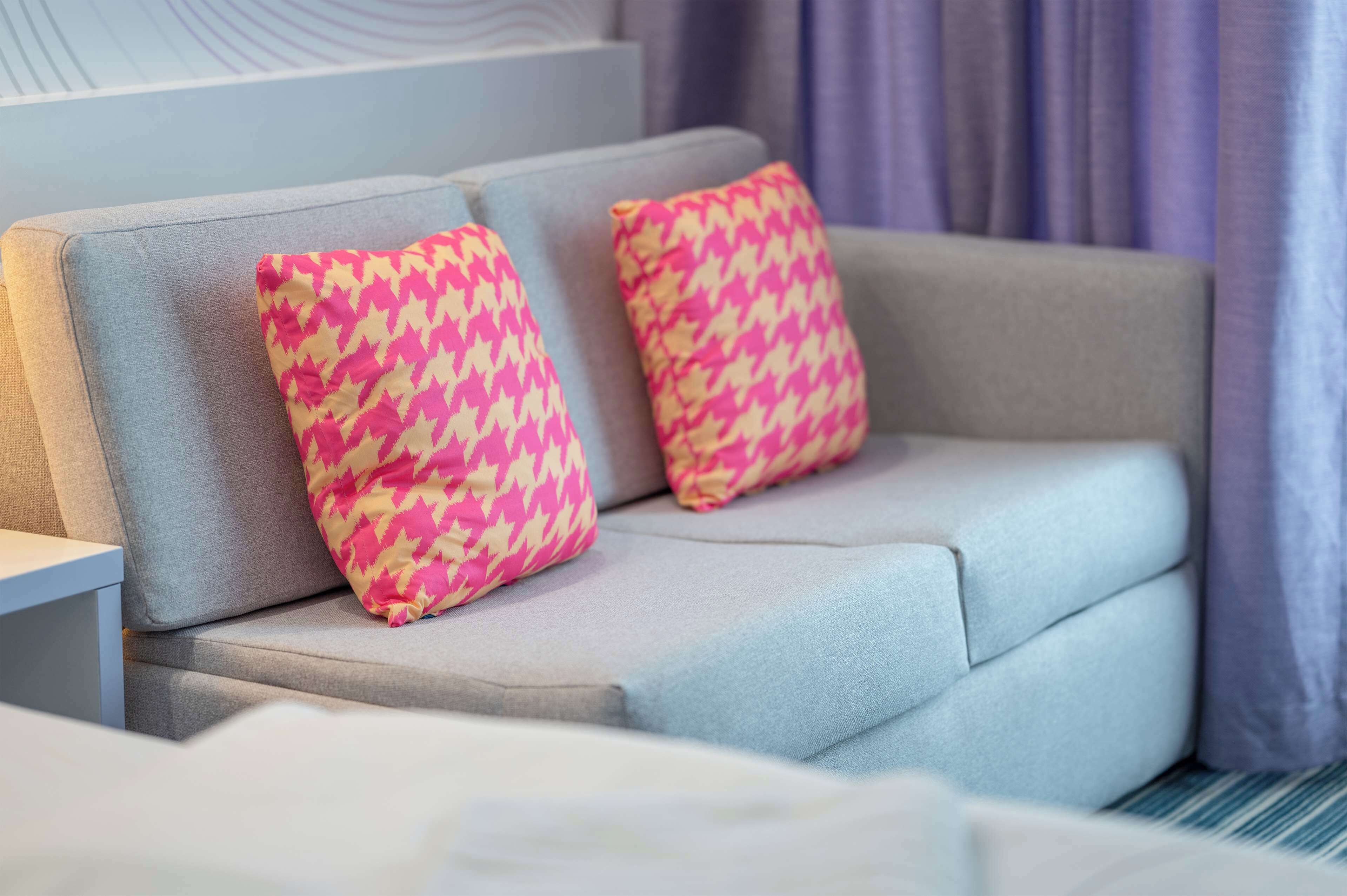 Um sofá moderno com duas almofadas estampadas, uma mesa lateral e vista para uma janela com cortinas.