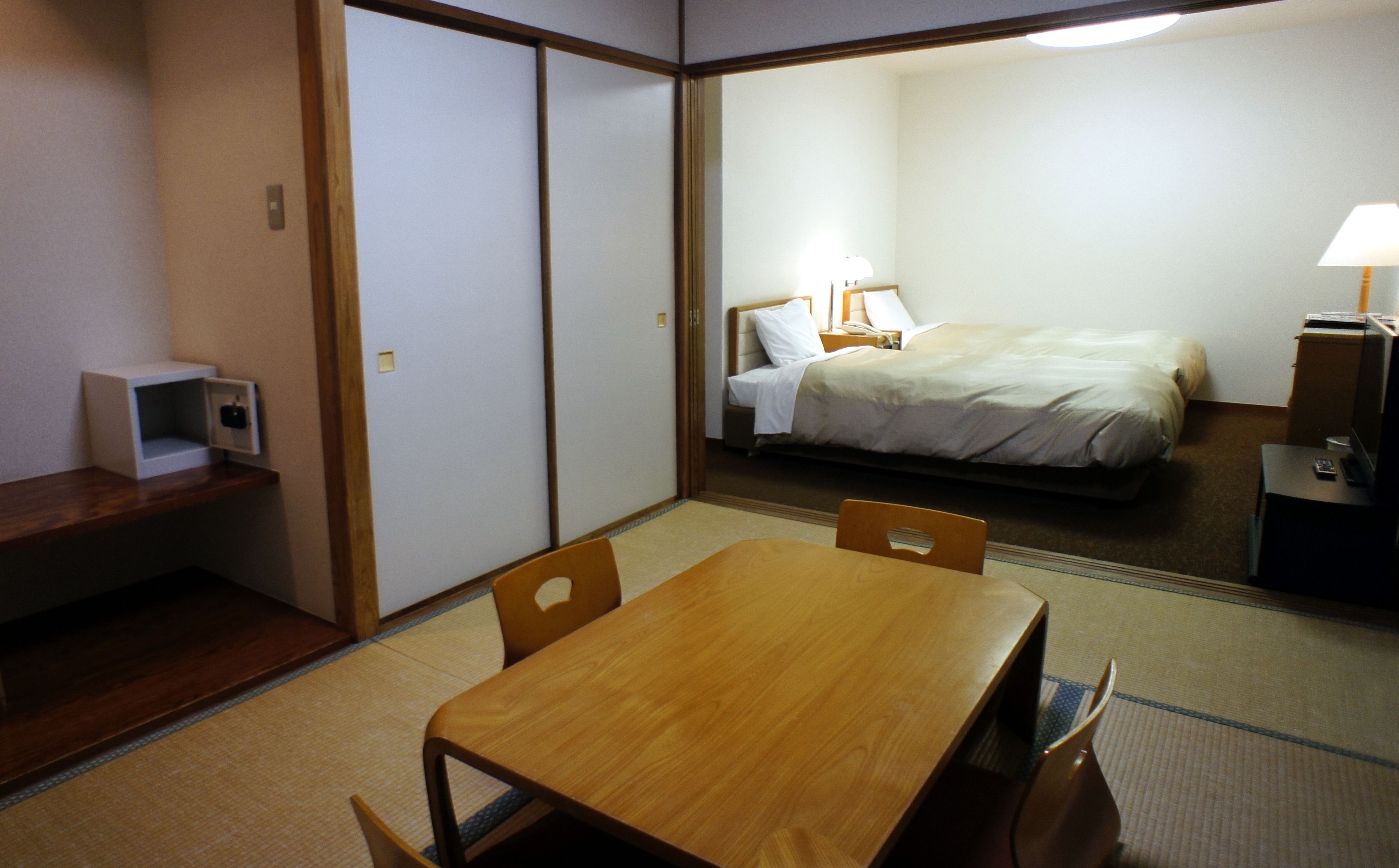 Quarto de hotel com mesa e cadeiras de madeira, cama com roupa de cama branca, luminária de cabeceira e uma pequena TV sobre um suporte.
