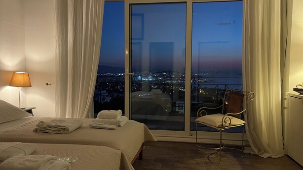 Quarto de hotel com uma grande porta de correr de vidro para o exterior, uma cama com toalhas dobradas, uma cadeira e uma luminária.
