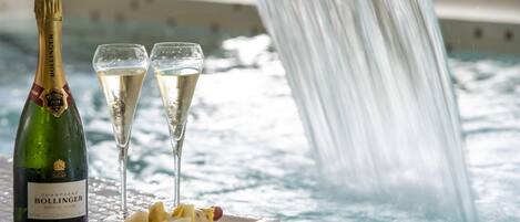 Uma garrafa de champanhe Bollinger, duas taças de champanhe e um prato com frutas sobre uma mesa perto de uma banheira de hidromassagem com cachoeira.