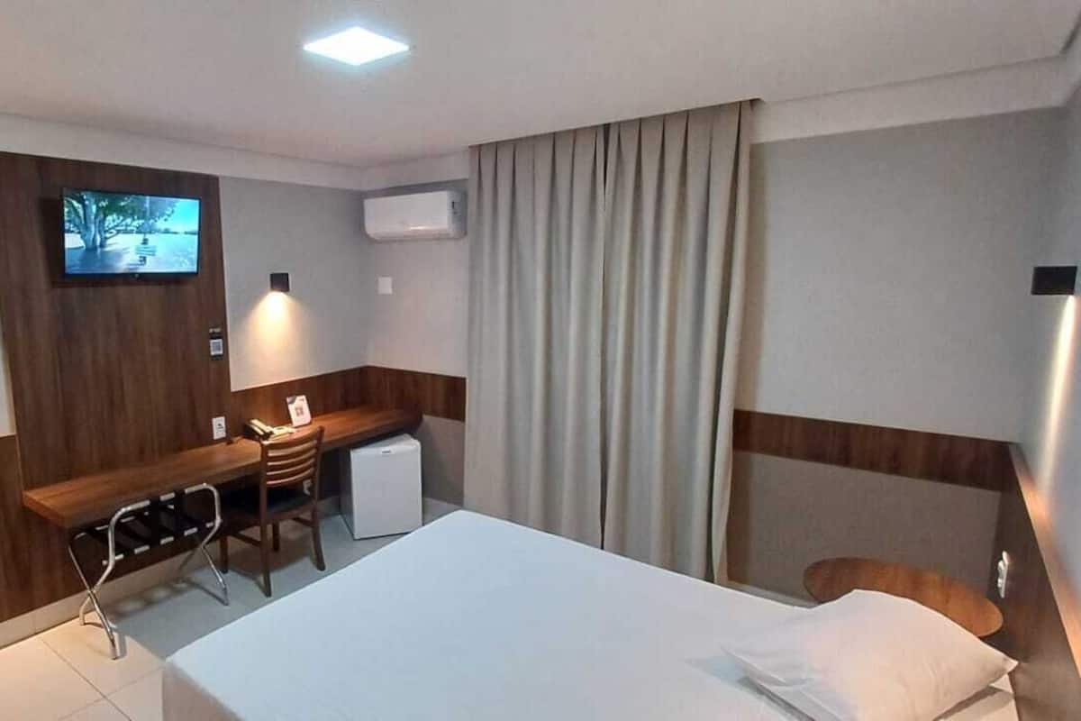 Quarto de hotel com uma cama, uma escrivaninha com cadeira, uma televisão na parede e uma janela com cortinas.