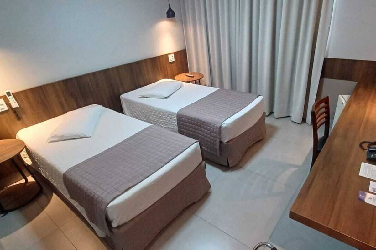 Quarto de hotel com duas camas, uma secretária de madeira, uma cadeira, uma mesinha e uma televisão.