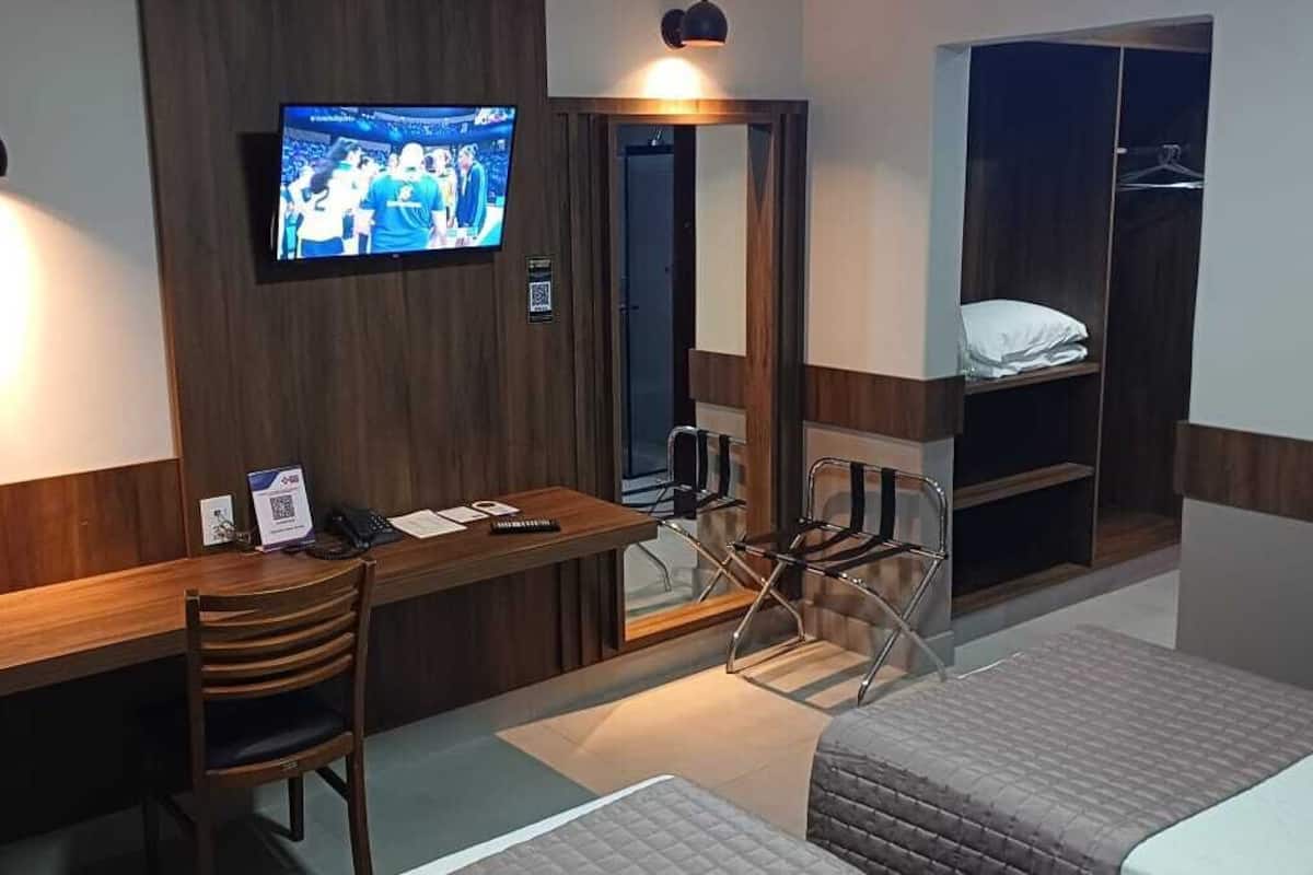 Quarto de hotel com escrivaninha de madeira, uma cadeira, cama, televisão fixada na parede e espelho.