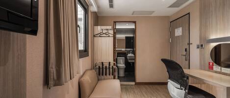 Quarto de hotel com cama, escrivaninha, poltrona, sofá, televisão e banheiro visível por uma porta aberta.