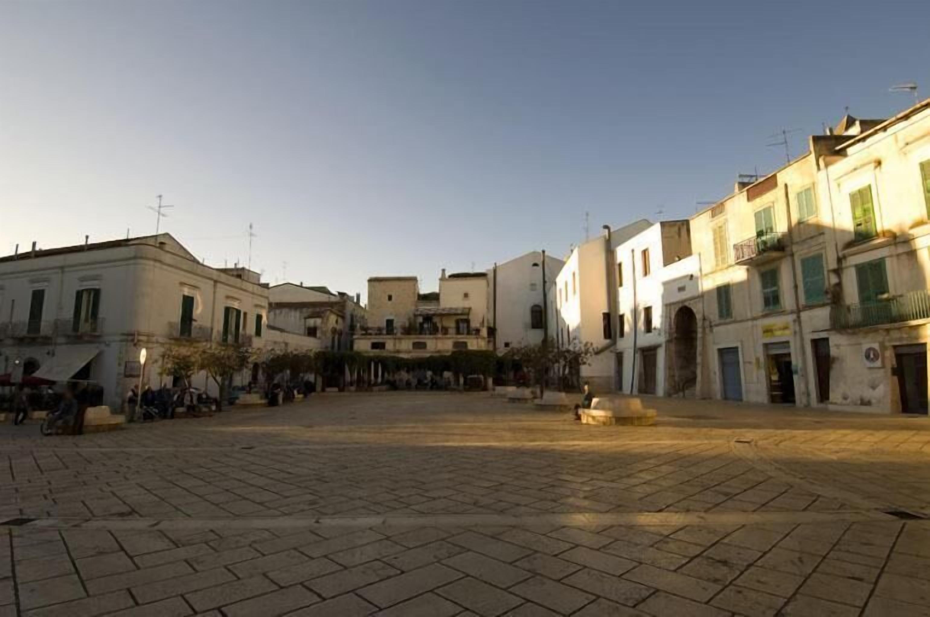 Uma praça pavimentada com edifícios em ambos os lados, apresentando um espaço aberto central e algumas árvores.