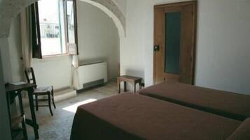 Quarto com duas camas, porta de madeira, uma pequena mesa, uma cadeira e janela com teto em arco.