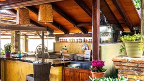 Uma área de bar ao ar livre rústica, com teto de madeira, cadeiras de vime, pia e diversos vasos de plantas.