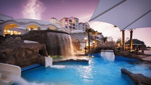 Piscina de hotel com cachoeira, palmeiras e um edifício com várias varandas ao fundo.