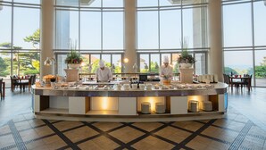 Espaço de buffet com chefs preparando alimentos, grandes janelas e vista para o exterior.