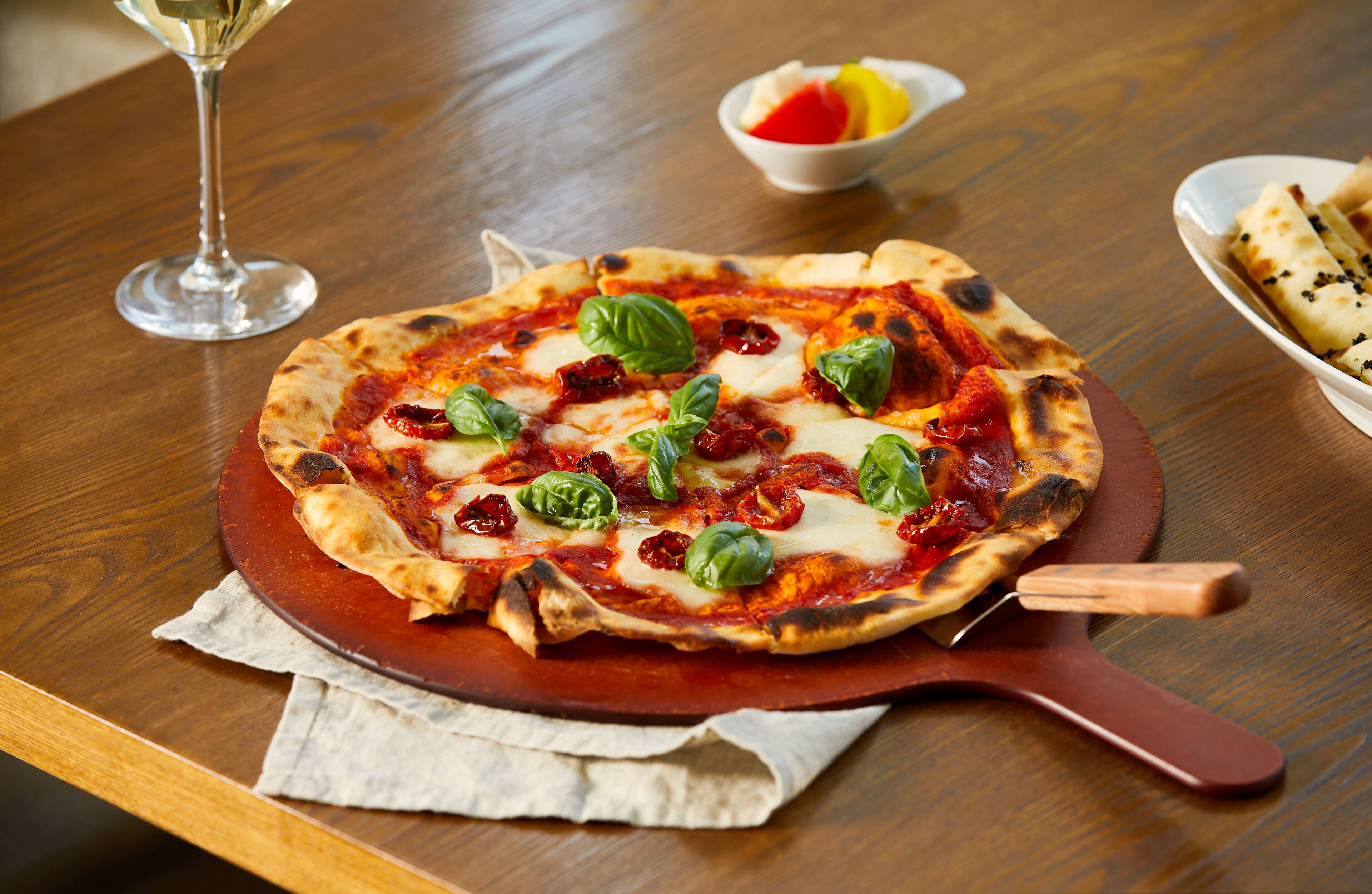 Pizza sobre uma mesa de madeira, com uma taça de vinho e uma tigela de frutas ao fundo.