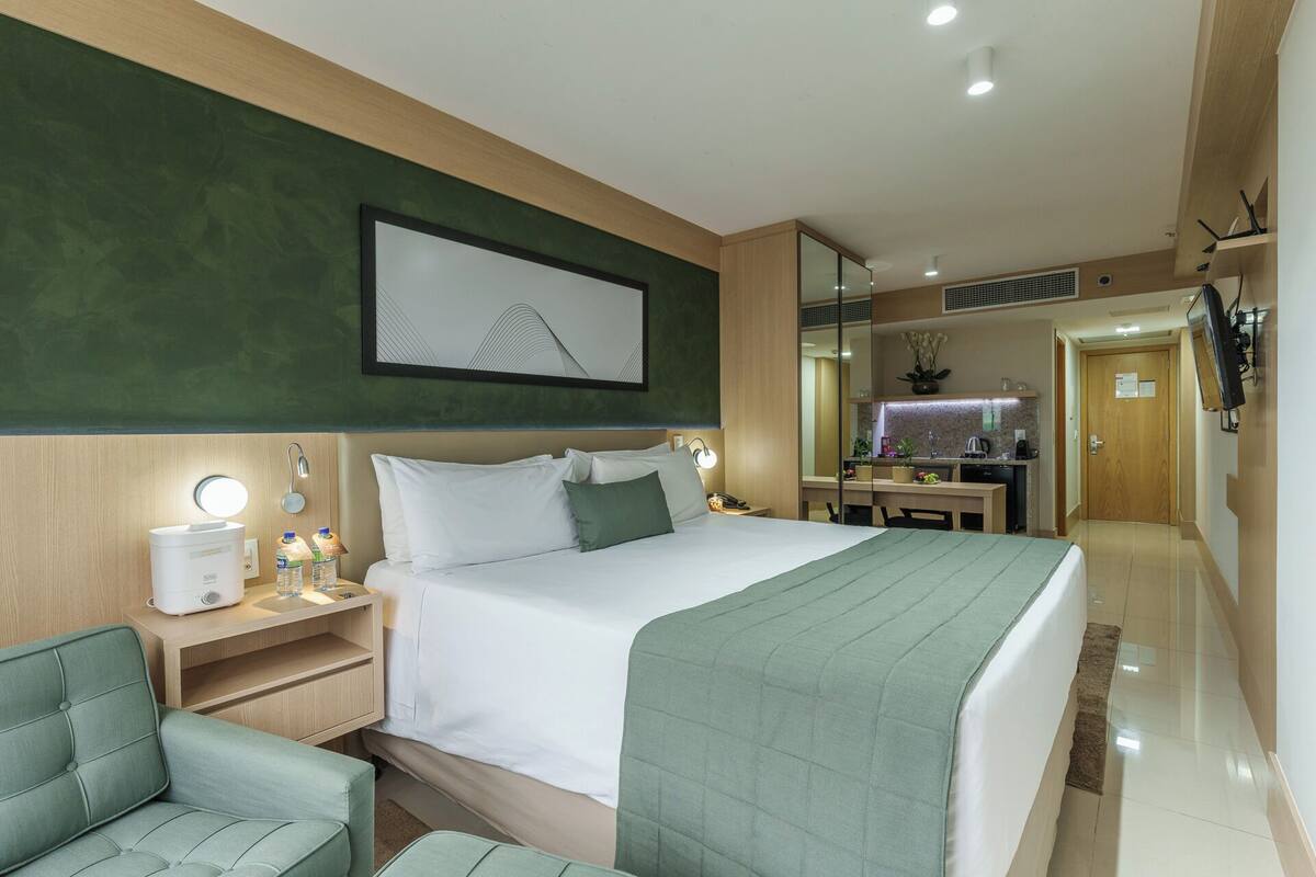Quarto de hotel com cama, mesas de cabeceira, escrivaninha, cadeira, espelho e cozinha compacta.