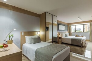 Quarto de hotel com duas camas, uma escrivaninha com abajur, um espelho e uma janela com vista para edifícios.