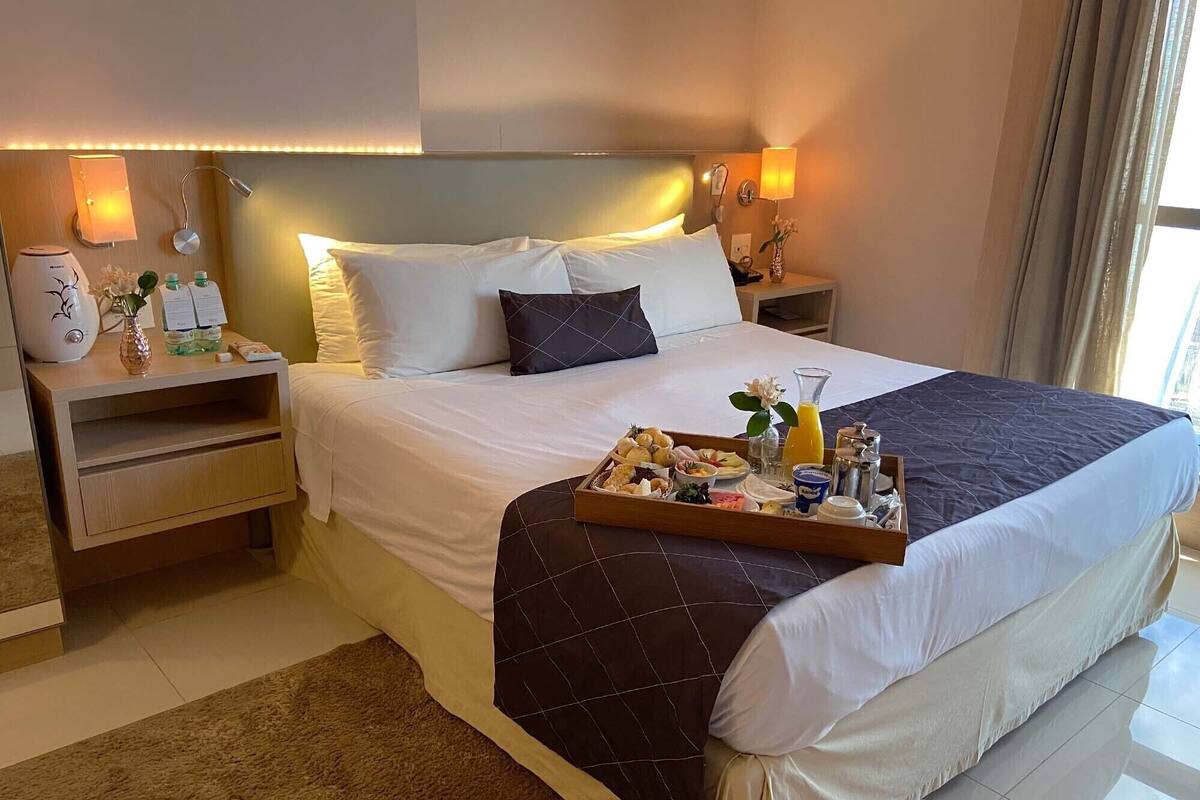 Quarto de hotel com uma cama grande, bandeja de itens de café da manhã, mesa de cabeceira com abajur e mesa lateral com vaso e telefone.