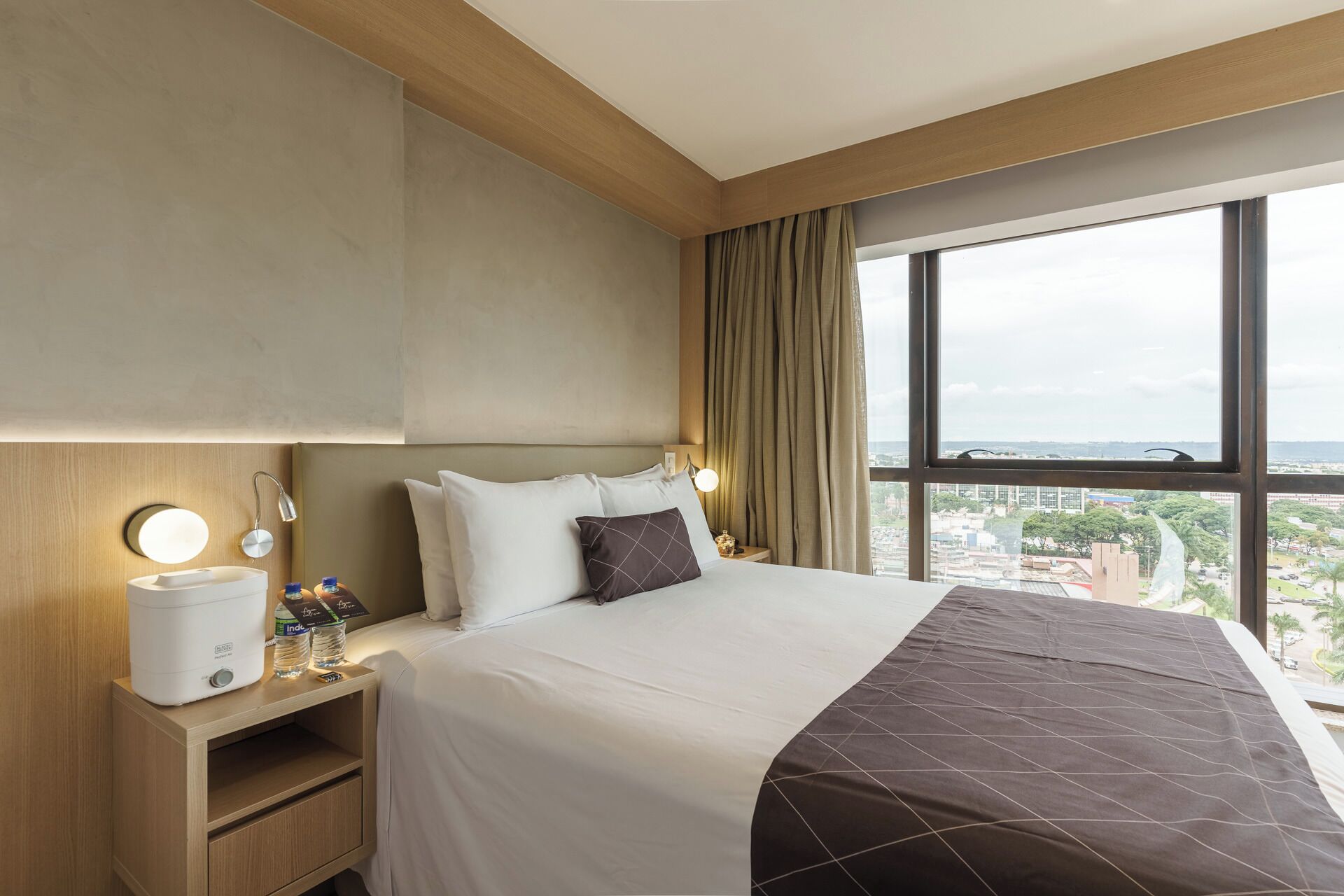 Quarto de hotel com uma cama grande, uma mesa de cabeceira com abajur, um dispensador de água e vista para a cidade através de amplas janelas.