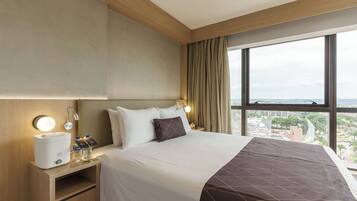 Quarto de hotel com uma cama grande, uma mesa de cabeceira com abajur, um dispensador de água e vista para a cidade através de amplas janelas.