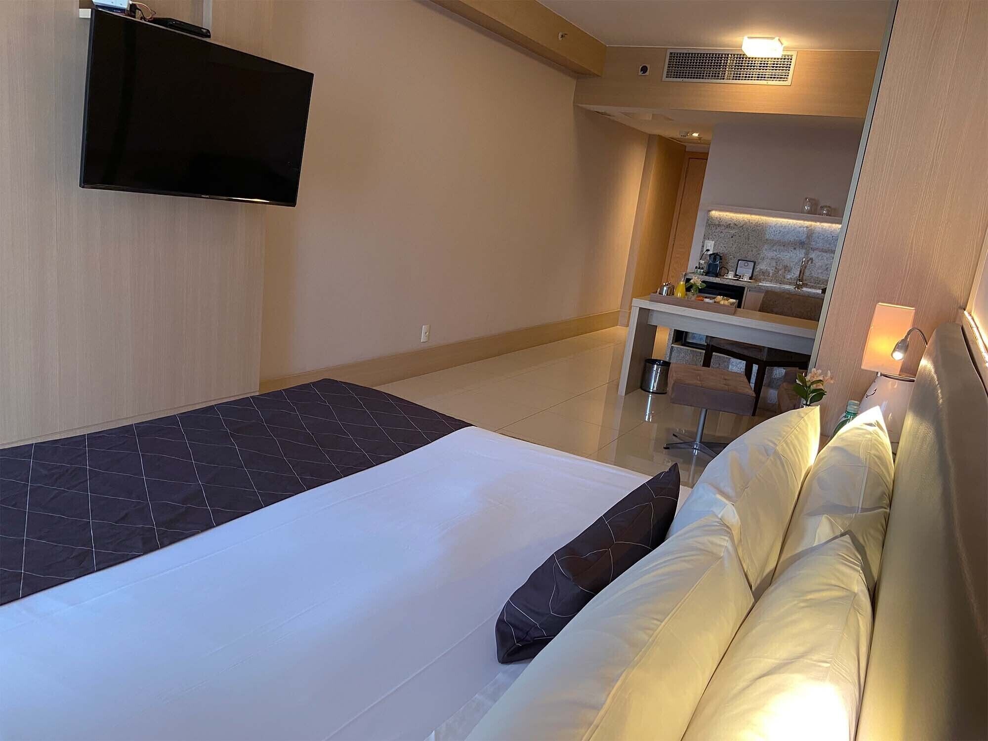 Quarto de hotel com cama, televisão na parede, pequeno espaço com cozinha e banheiro ao fundo.