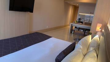 Quarto de hotel com cama, televisão na parede, pequeno espaço com cozinha e banheiro ao fundo.