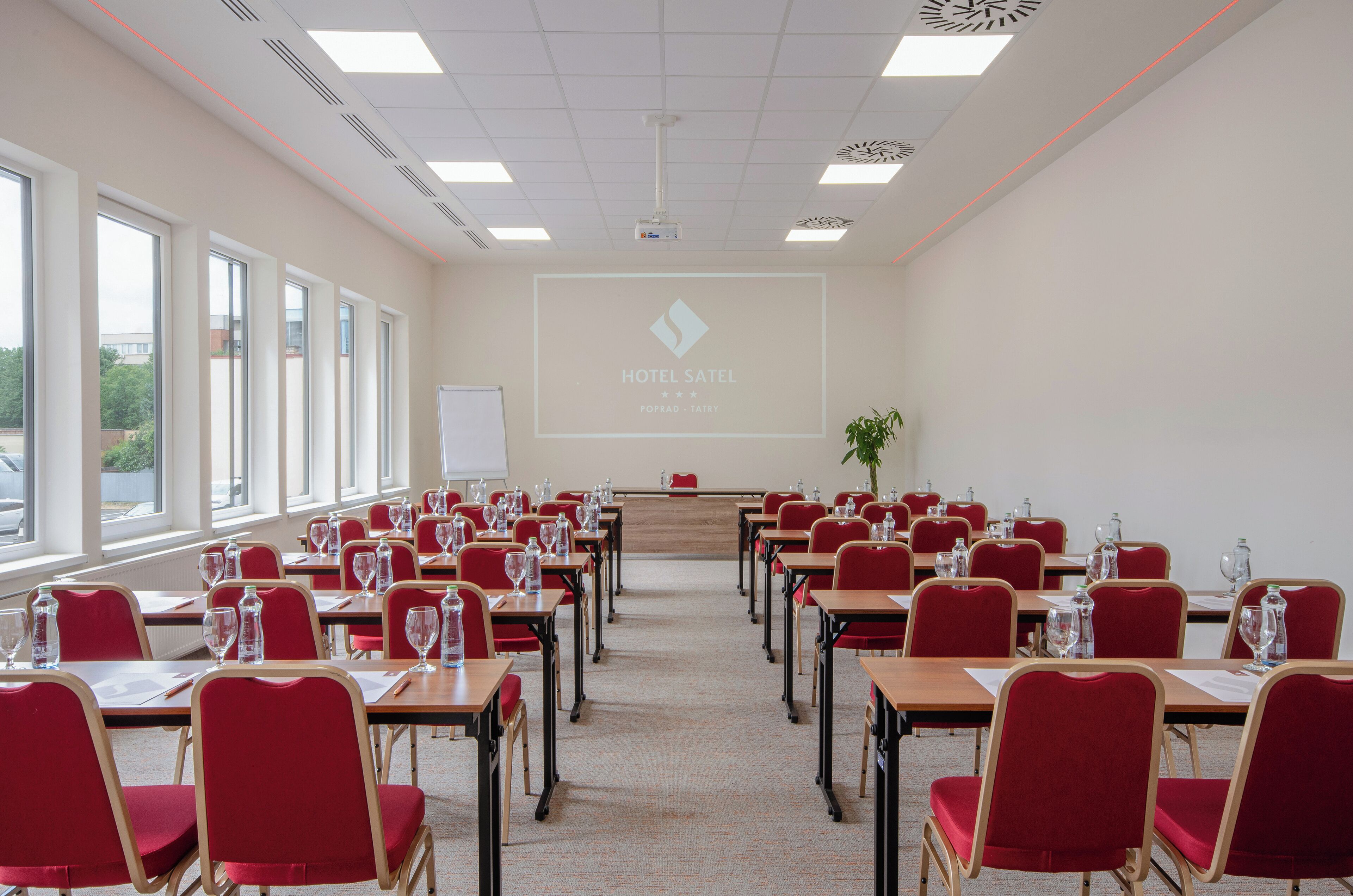 Uma sala de conferências com fileiras de mesas e cadeiras, um quadro branco e uma tela de projetor exibindo 'HOTEL SATEL POPRAD - ENTRADA'.