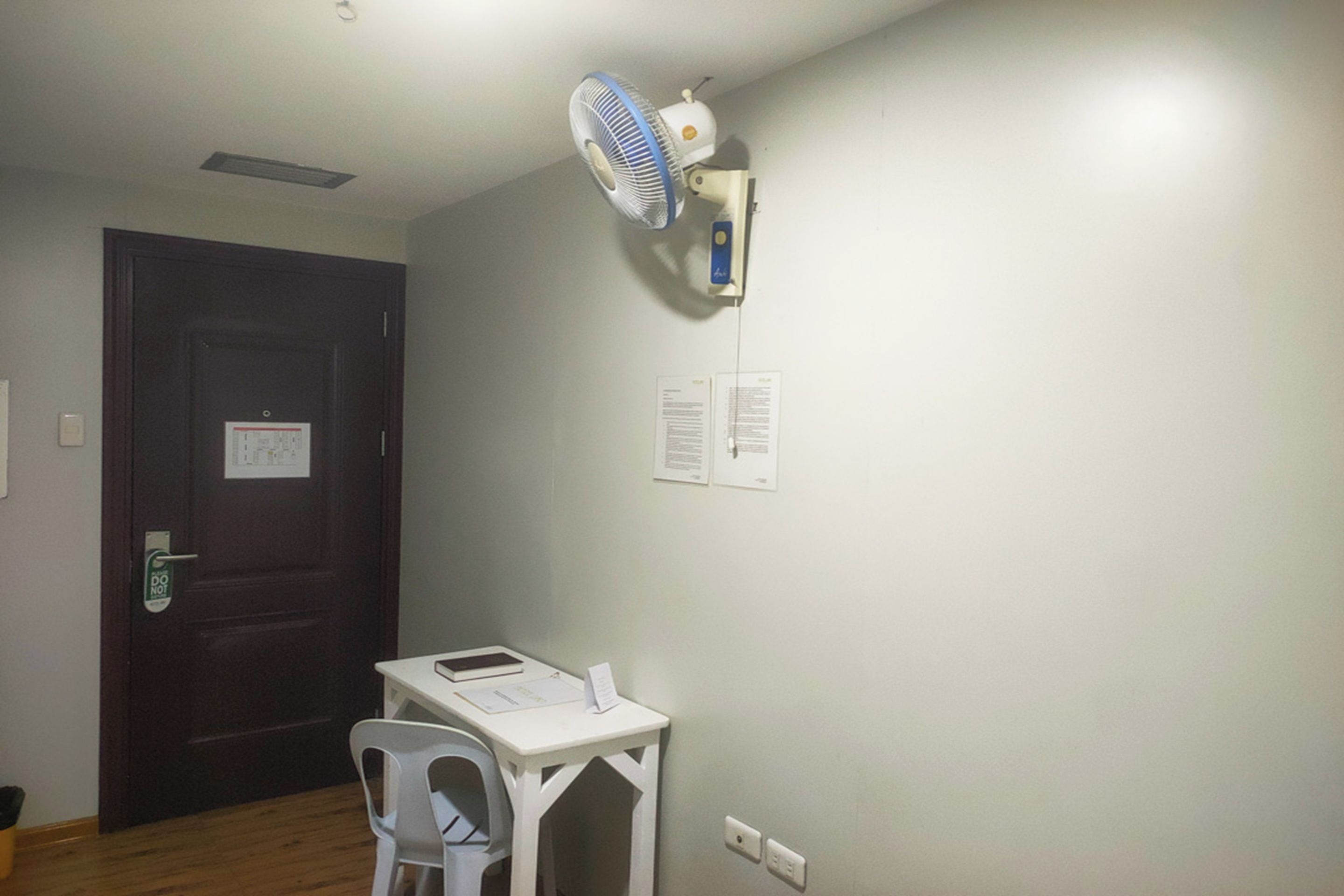 Quarto com escrivaninha, cadeira, ventoinha na parede e porta com placa.
