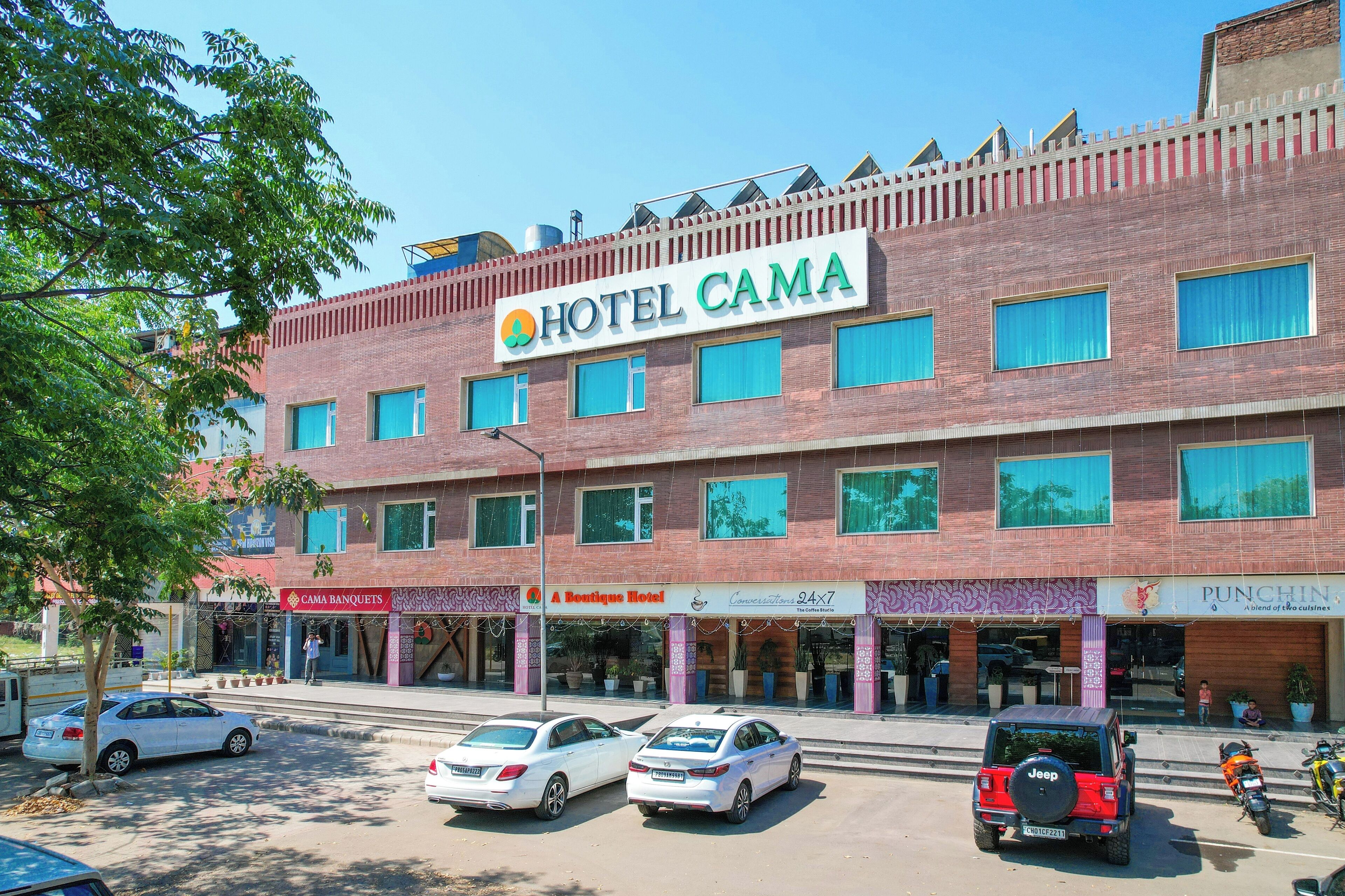 Um hotel chamado "Hotel Cama", com vários andares, fachada de tijolos vermelhos e diversas lojas no nível da rua, incluindo "Cama Banquets" e "A Boutique Hotel".