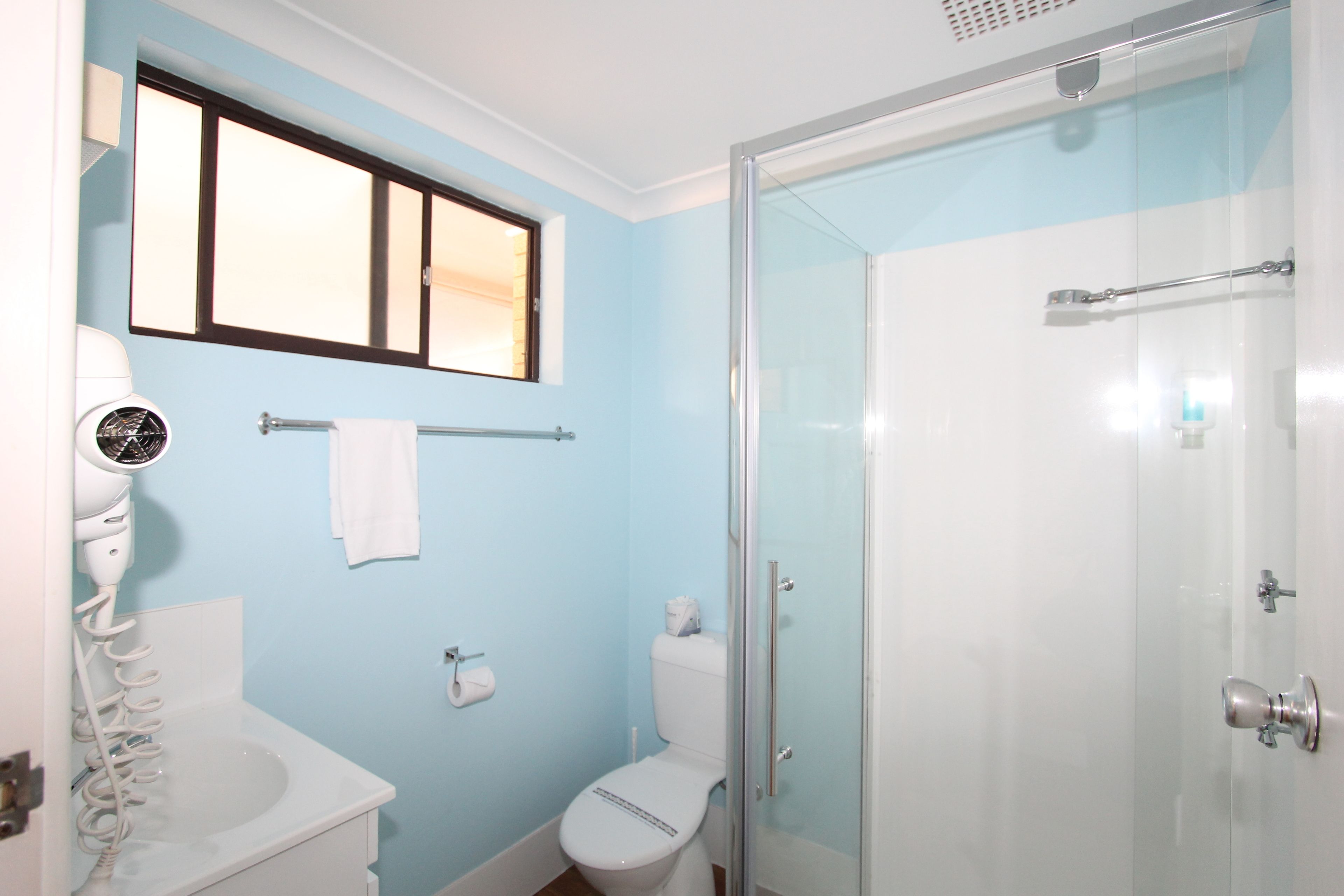 Uma casa de banho com chuveiro de porta de vidro, pia branca, sanita e secador de cabelo.
