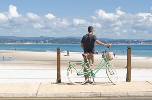 Um homem parado ao lado de uma bicicleta verde numa praia, com céu claro e montanhas ao longe.