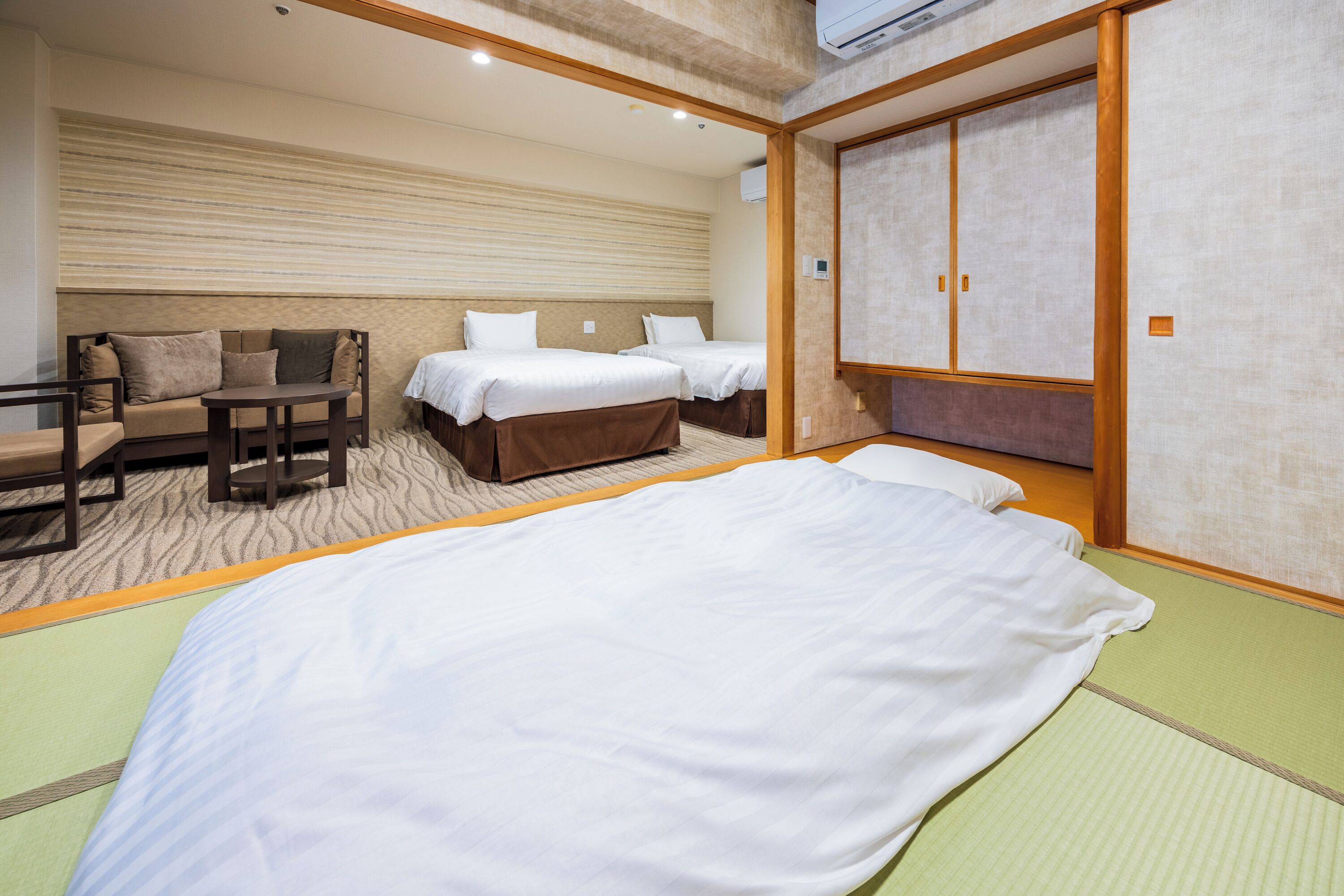 Quarto de hotel com duas camas, um sofá, uma mesa pequena e uma porta tradicional japonesa deslizante.