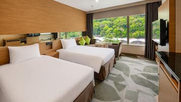 Quarto de hotel com duas camas, escrivaninha, cadeira, televisão e uma grande janela com vista para o verde.
