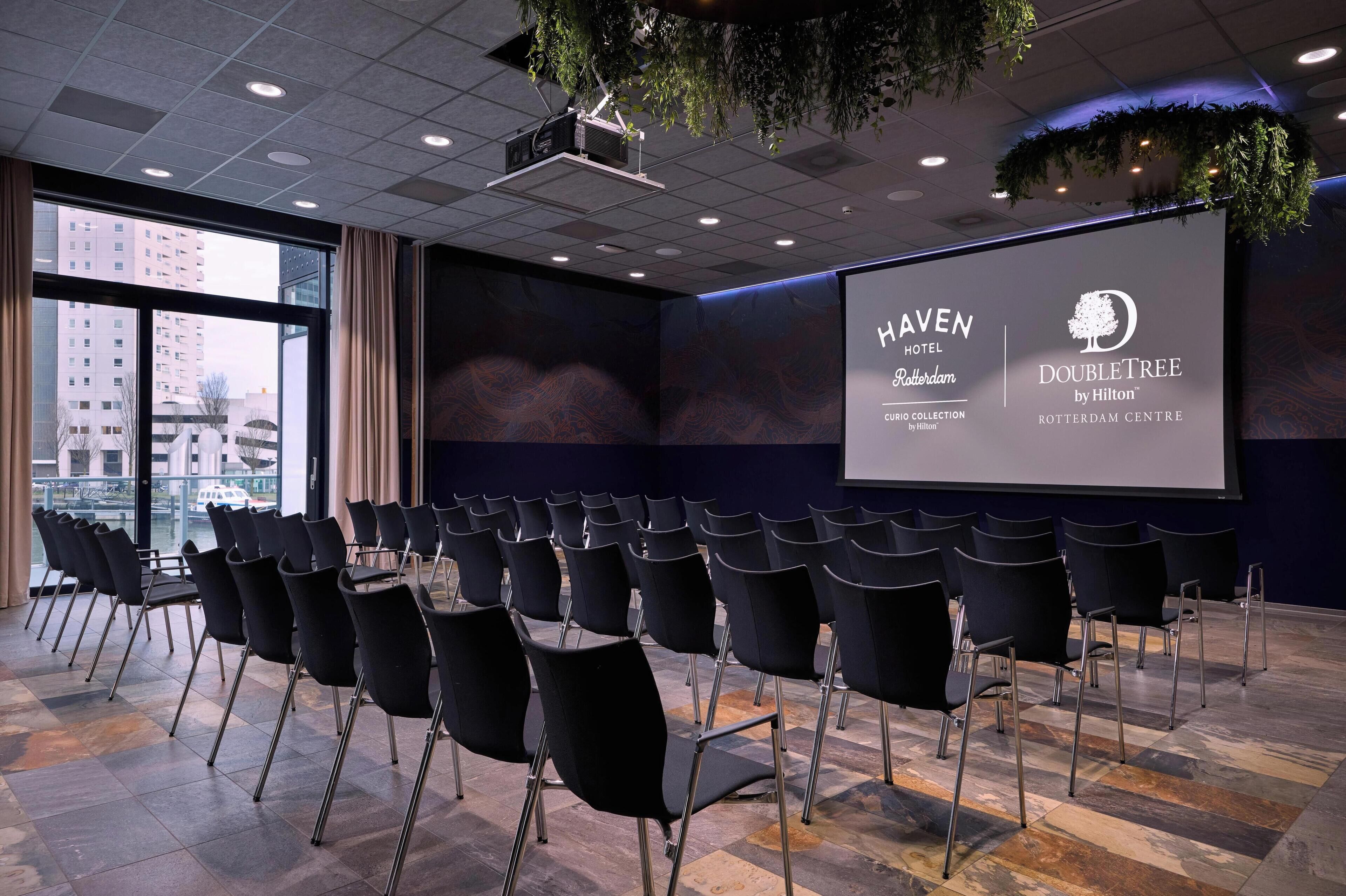 Uma sala de conferências com fileiras de cadeiras pretas, uma tela grande exibindo o nome do hotel e vista para prédios através de amplas janelas.