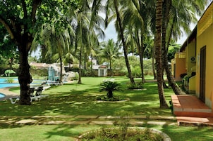Um jardim tropical com palmeiras, uma piscina e um edifício com fachada amarela.