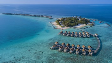 Resort de bangalôs sobre a água com um píer que liga à ilha principal, águas turquesa cristalinas e uma linha de costa distante.