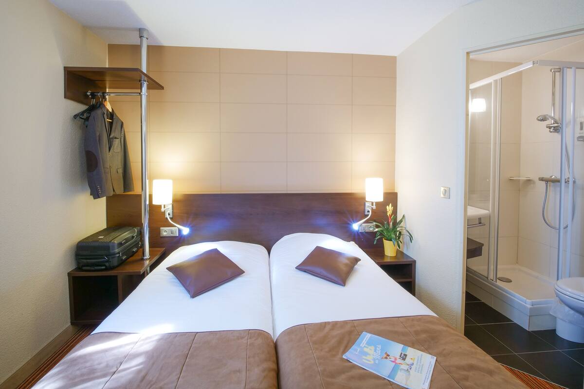 Quarto de hotel com duas camas, chuveiro, mesa de cabeceira com abajur, cabideiro e mala.
