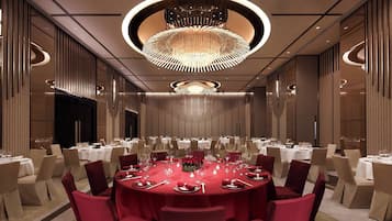 Uma sala de jantar formal com mesas redondas, cadeiras estofadas em vermelho e um grande candelabro.