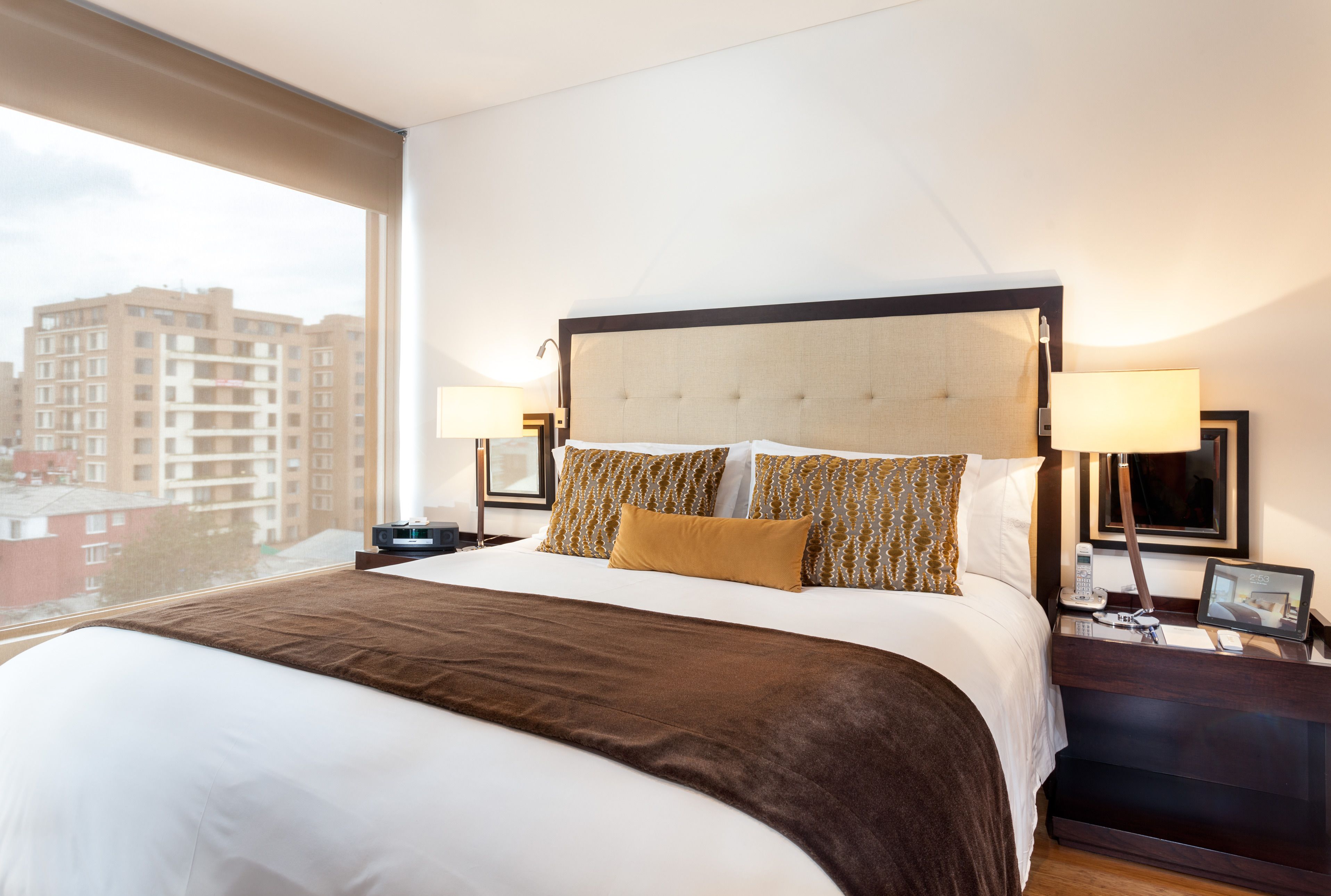 Quarto de hotel com uma cama grande, duas mesas de cabeceira com luminárias, vista de edifícios por uma janela ampla e uma paisagem urbana.