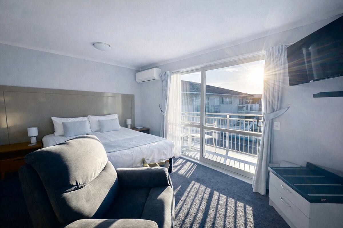 Quarto de hotel com cama, poltrona, escrivaninha, televisão e varanda com vista para a cidade.