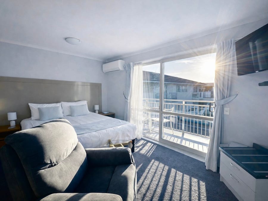 Quarto de hotel com cama, poltrona, escrivaninha, televisão e varanda com vista para a cidade.