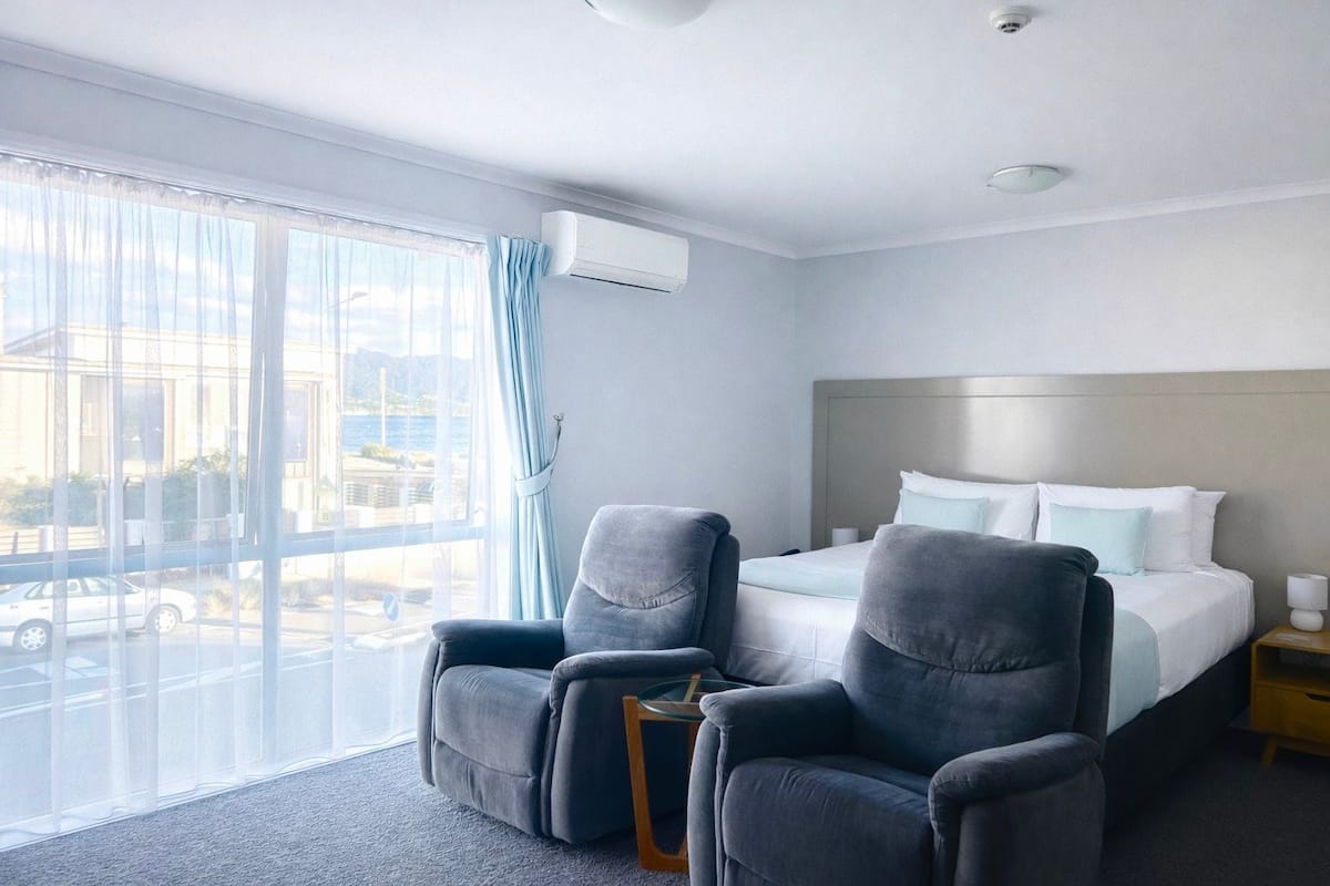Quarto de hotel com uma cama, duas poltronas, uma mesa de cabeceira e uma janela grande com cortinas.