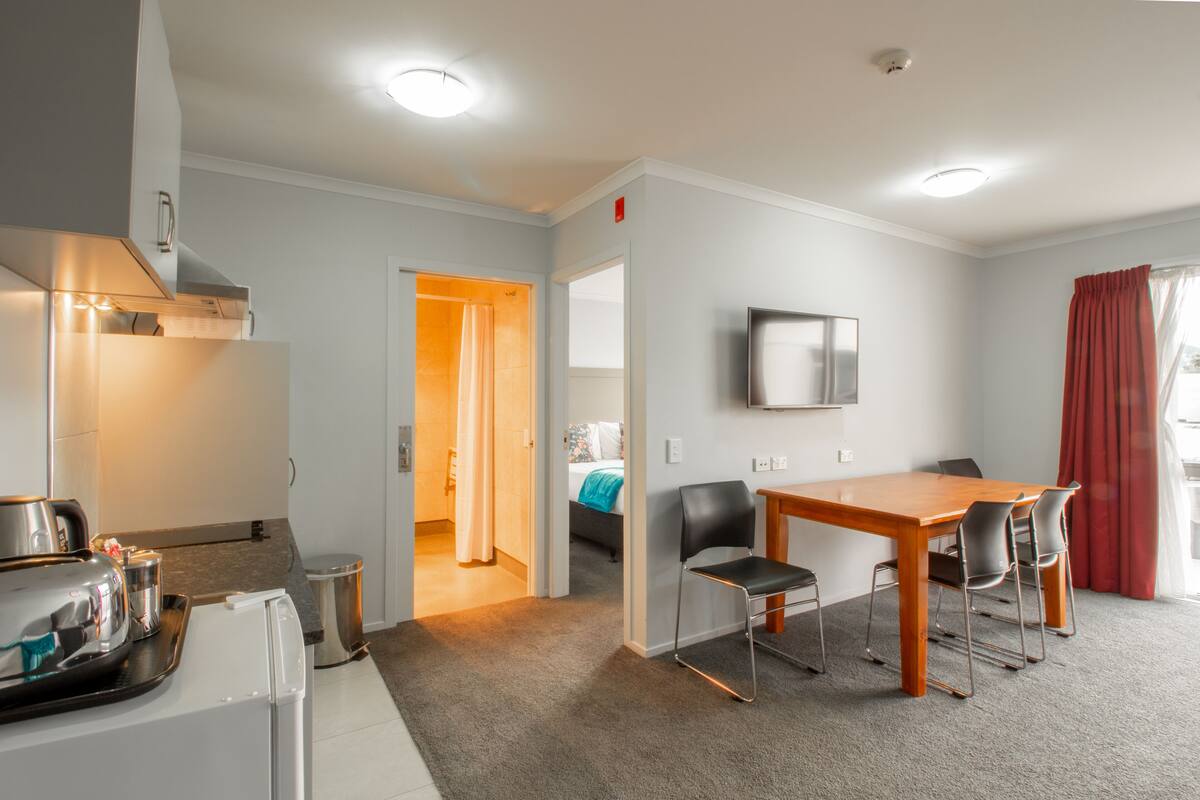 Quarto de hotel moderno com cozinha compacta, área de refeições com mesa e cadeiras, televisão presa na parede e cortina vermelha.
