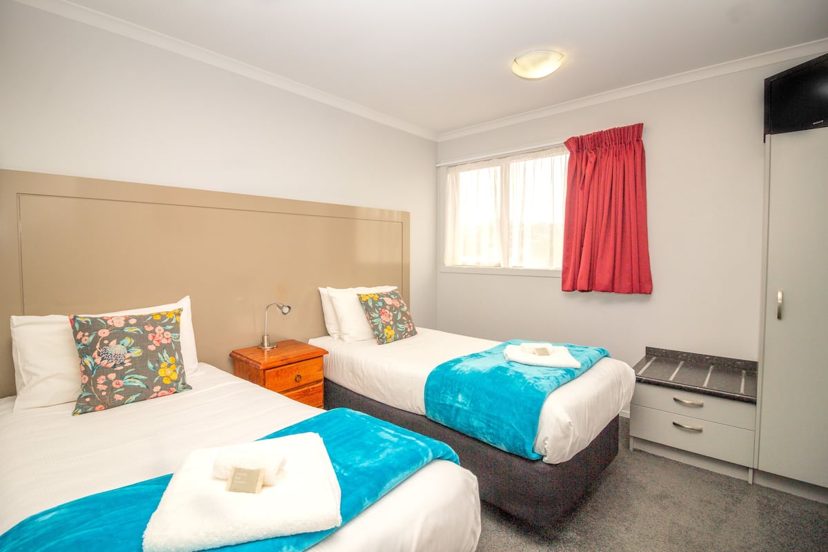 Quarto de hotel com duas camas, uma janela com cortinas vermelhas, um guarda-roupa e uma pequena cozinha compacta.