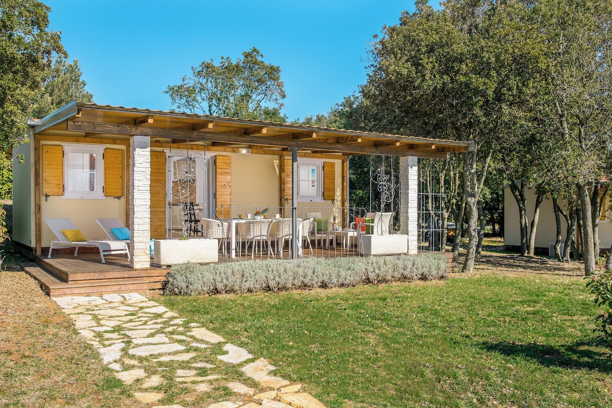 Um pátio externo aconchegante com pérgola de madeira, um caminho de pedra e área de estar com cadeiras e almofadas brancas.