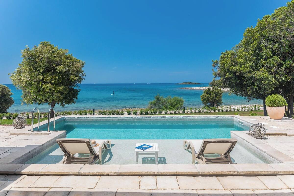 Piscina com vista para o mar, rodeada por árvores e uma pequena mesa com cadeiras.