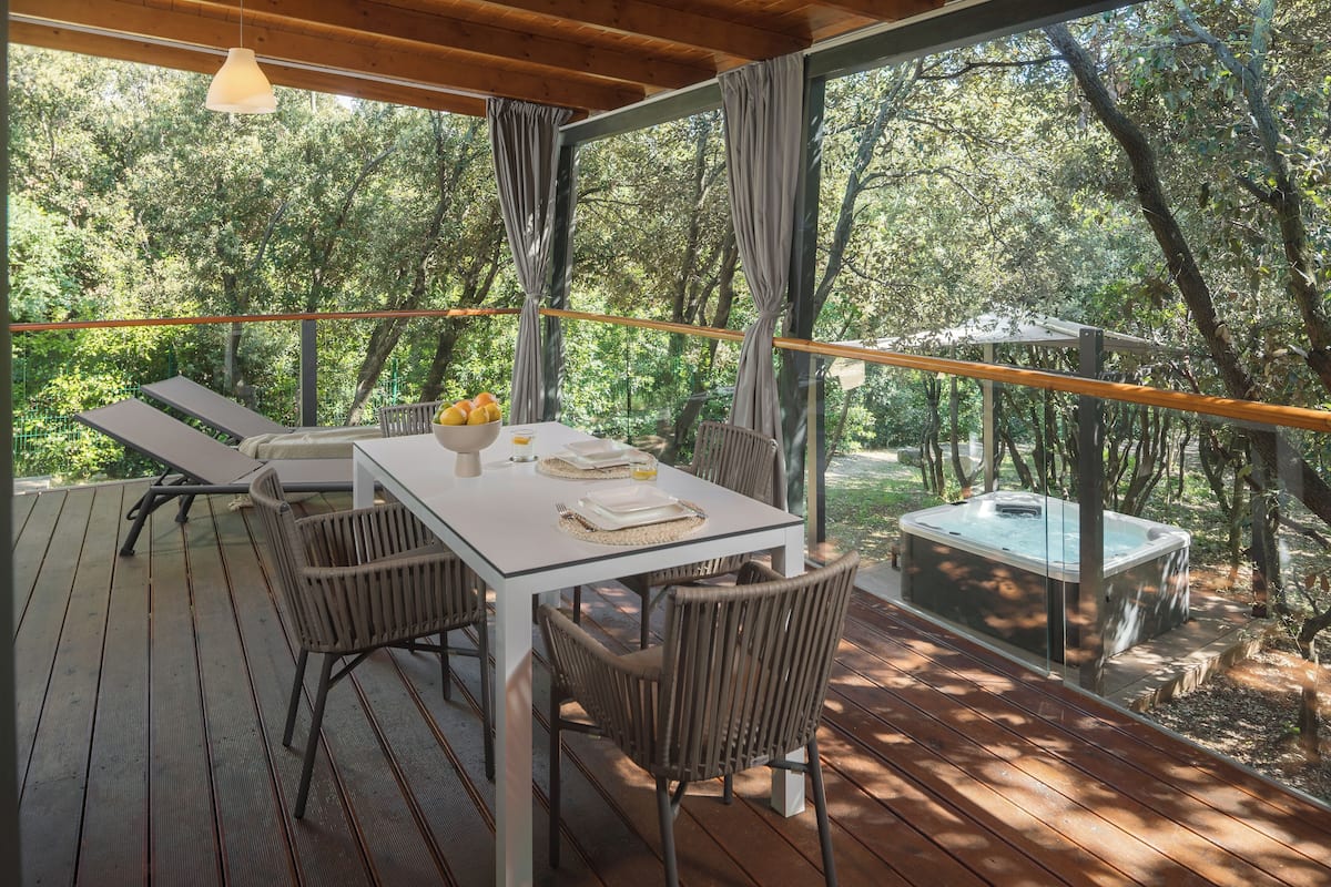 Uma área de refeições ao ar livre coberta, com uma mesa posta para dois, rodeada por cadeiras de vime, com vista para uma área arborizada.