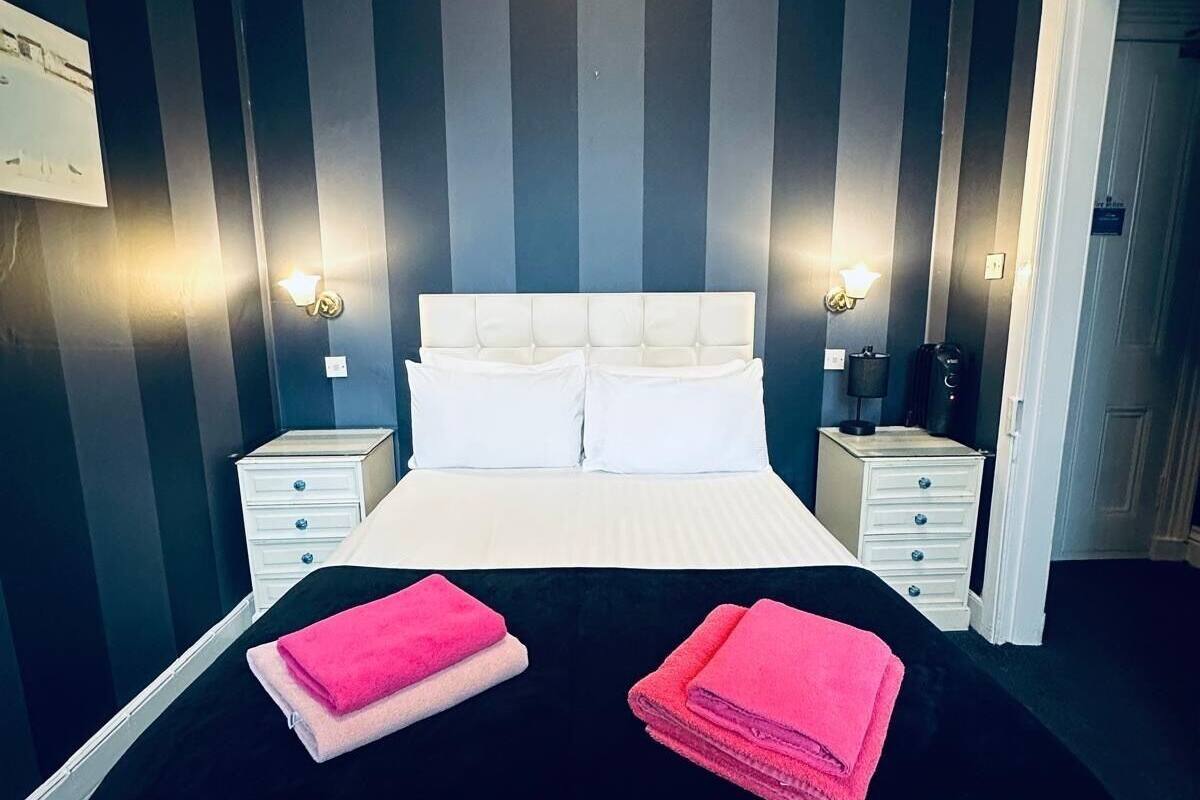 Quarto de hotel com uma cama grande, duas mesas de cabeceira, uma luminária de parede e uma porta que dá para outro cômodo.