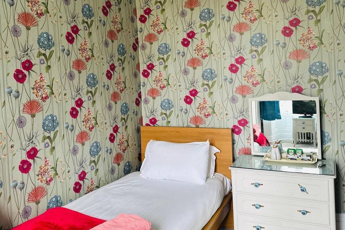 Um quarto com papel de parede floral, uma cama de madeira, uma cômoda branca e um espelho.