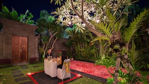 Uma área de refeições ao ar livre com uma mesa posta para dois, rodeada por vegetação exuberante e um canteiro de flores vermelhas.