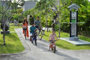 Uma trilha com uma família andando de bicicleta, cercada por vegetação e uma construção moderna com painéis informativos.