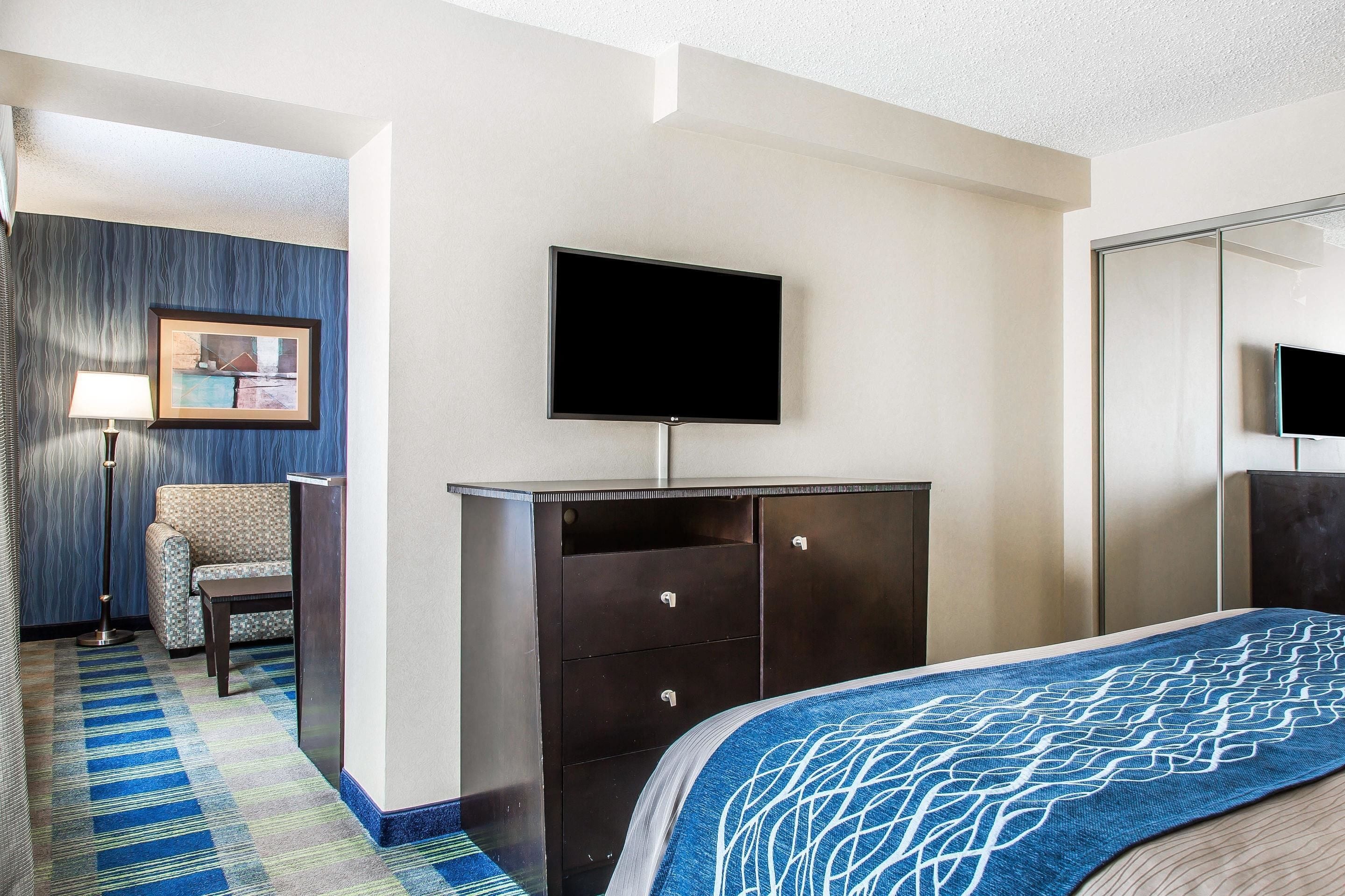 Quarto de hotel com cama, televisão de tela plana na parede, escrivaninha com luminária, cadeira e guarda-roupa.