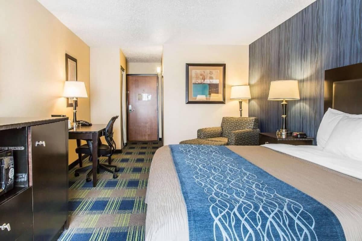 Quarto de hotel com cama, duas mesas de cabeceira com luminárias, escrivaninha com cadeira, porta e obra de arte na parede.