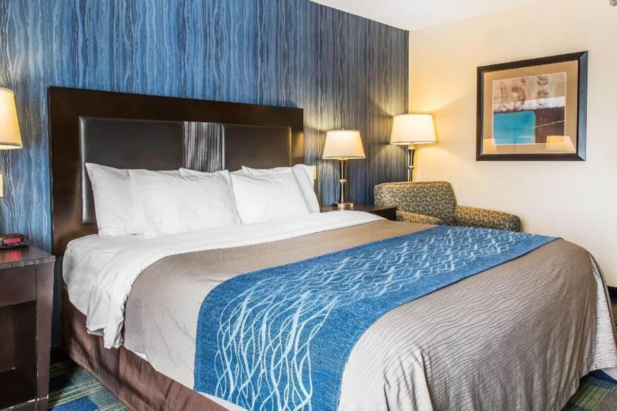 Quarto de hotel com uma cama grande, mesa de cabeceira, cadeira, abajur e um quadro na parede.