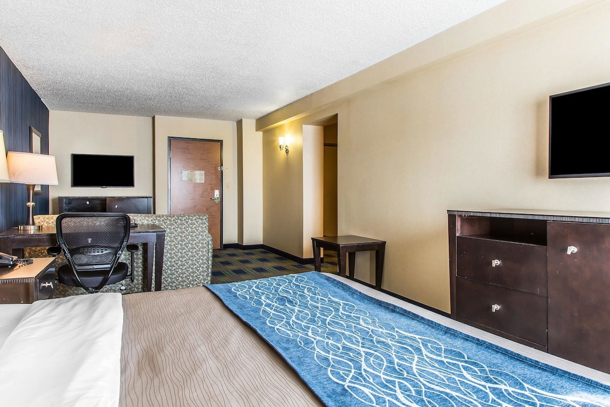 Quarto de hotel com uma cama, uma escrivaninha com luminária, uma cadeira, uma TV fixada na parede e um penteado.